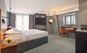 Quarto de hotel com cama, escrivaninha, cadeira e uma janela grande.