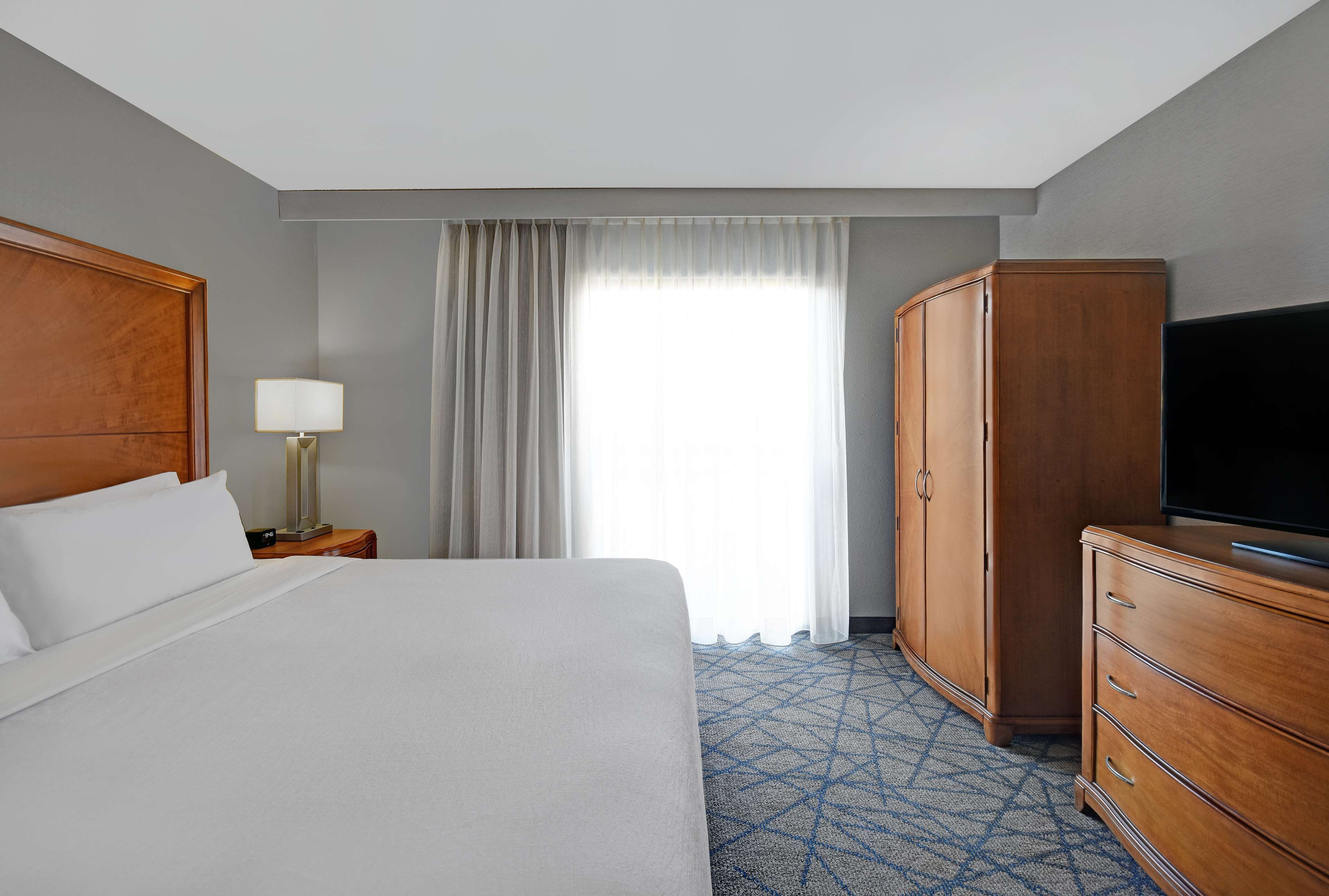 Quarto de hotel com uma cama grande, um guarda-roupa de madeira, uma televisão e uma mesa de cabeceira com abajur.