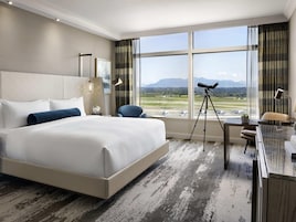 Quarto de hotel com uma cama grande, uma escrivaninha com abajur, uma cadeira, um telescópio e vista para uma pista de pouso e montanhas.