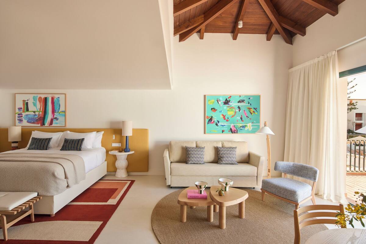 Quarto de hotel com cama, sofá, poltrona, mesa pequena e obras de arte nas paredes.