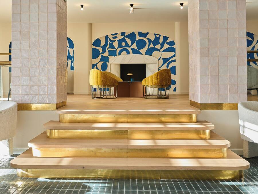 Um hall de hotel com uma escadaria de mármore, duas poltronas amarelas e uma parede decorativa com padrões azuis.