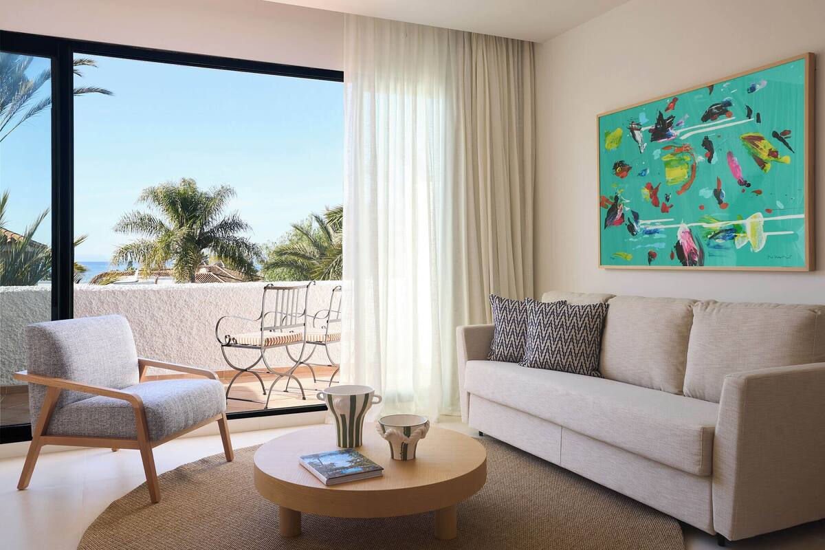 Sala de estar com sofá, poltrona e mesa de centro, um quadro na parede e vista para palmeiras e um prédio através de grandes janelas.