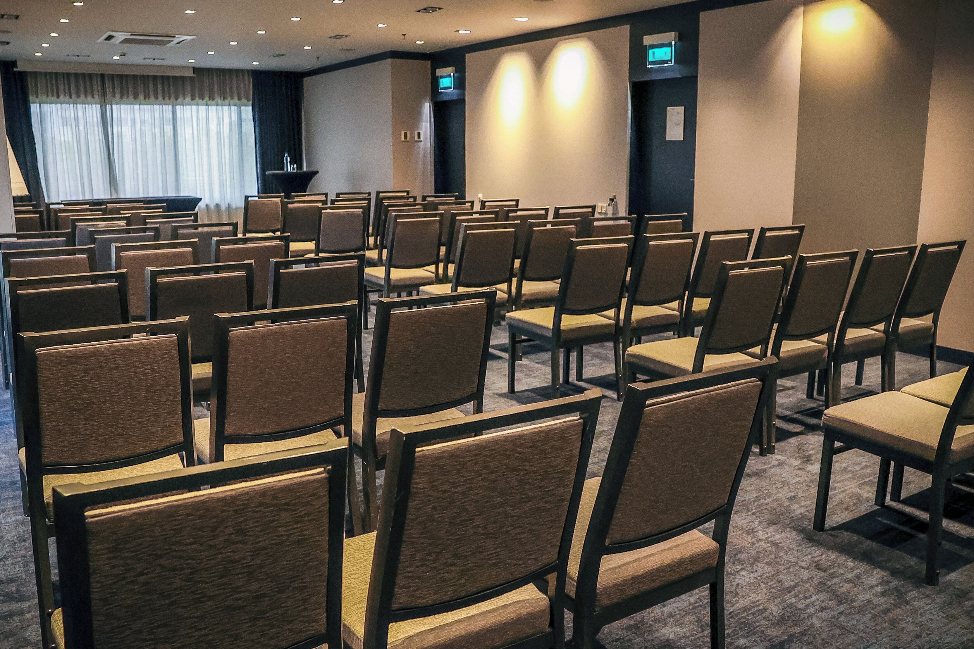 Sala de conferência com fileiras de cadeiras, um palanque e uma janela grande com cortinas.