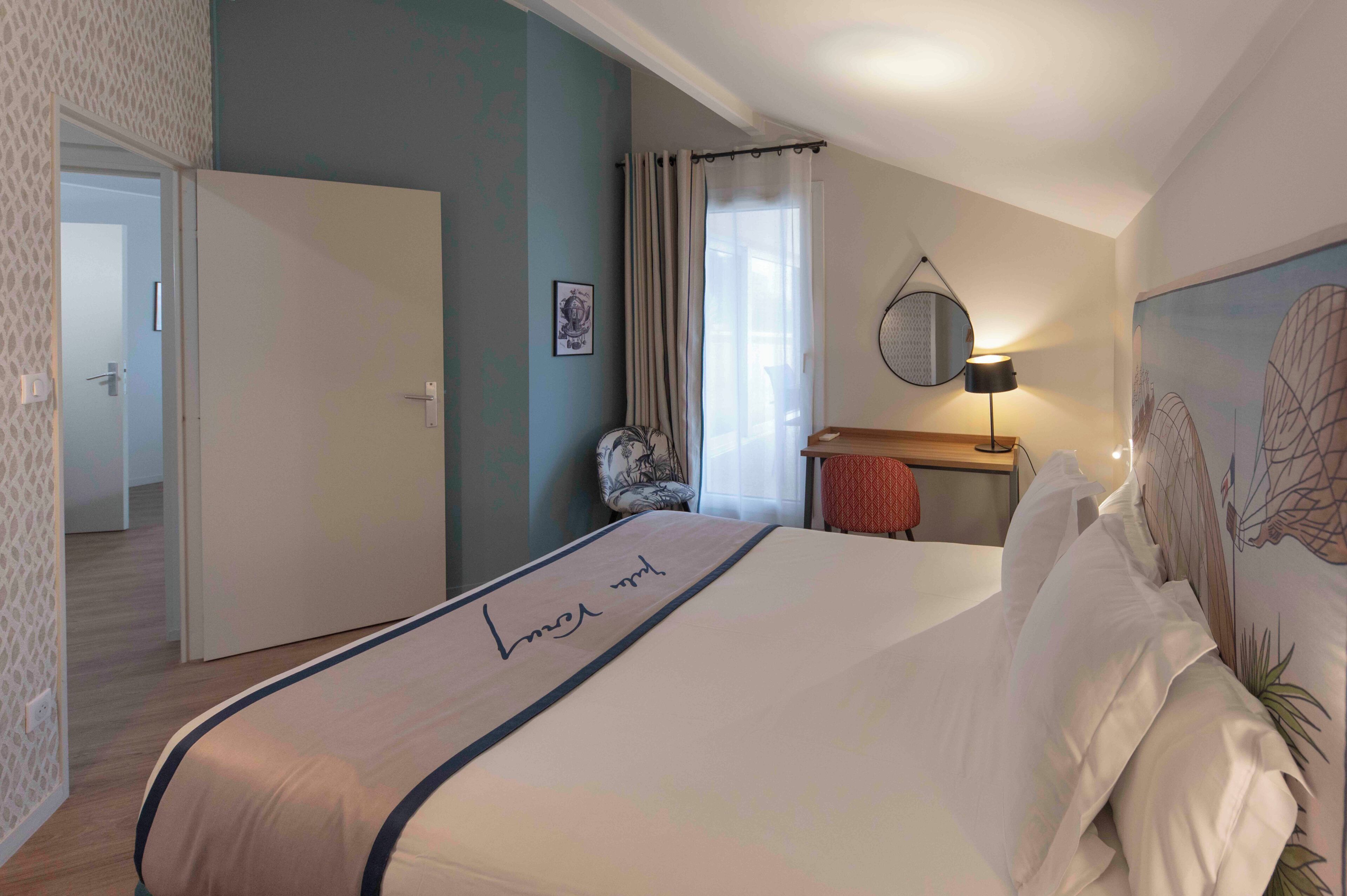 Quarto de hotel com cama, escrivaninha, cadeira, janela com cortinas e um quadro pendurado na parede.