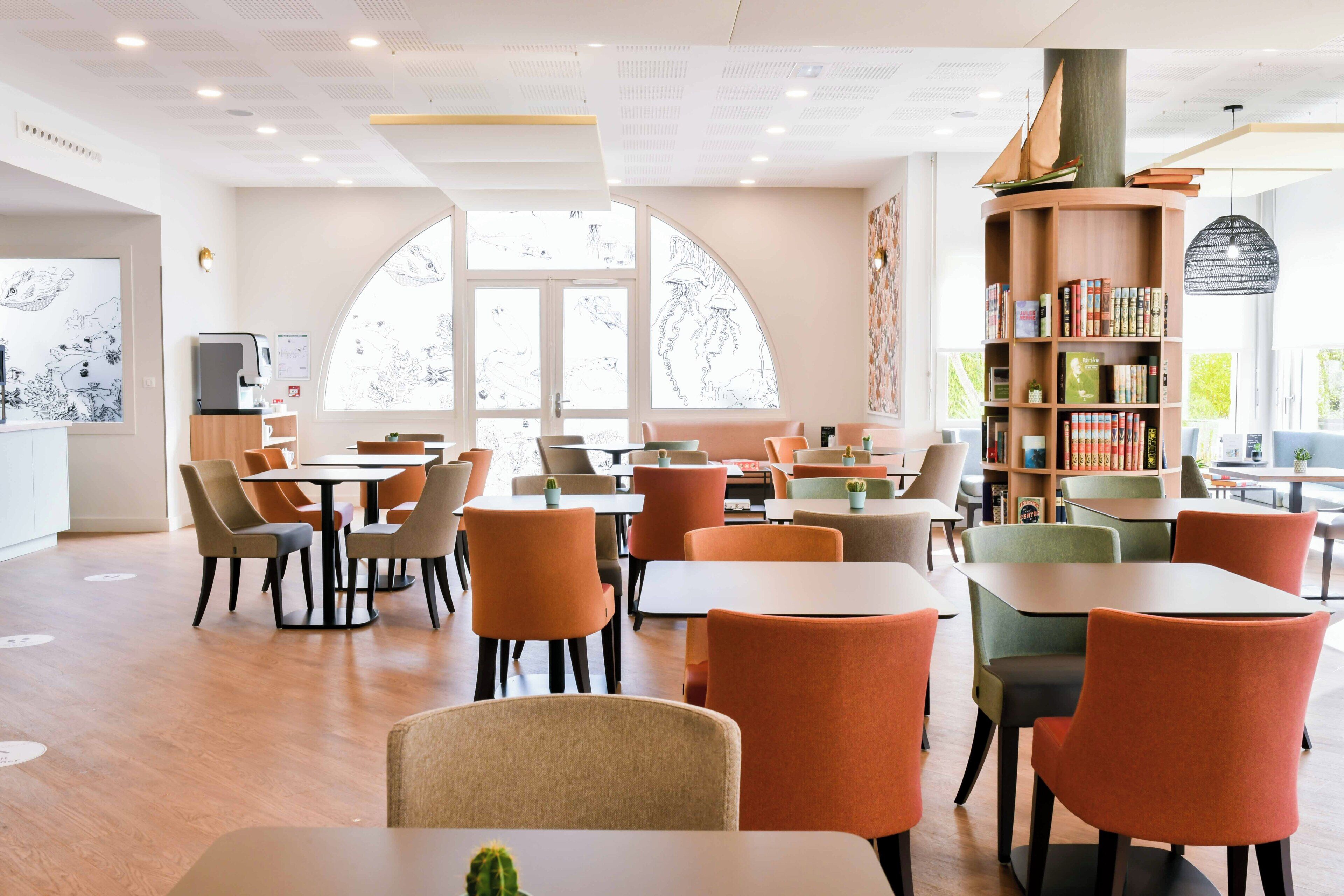Um café com várias mesas e cadeiras, uma estante de livros e grandes janelas.