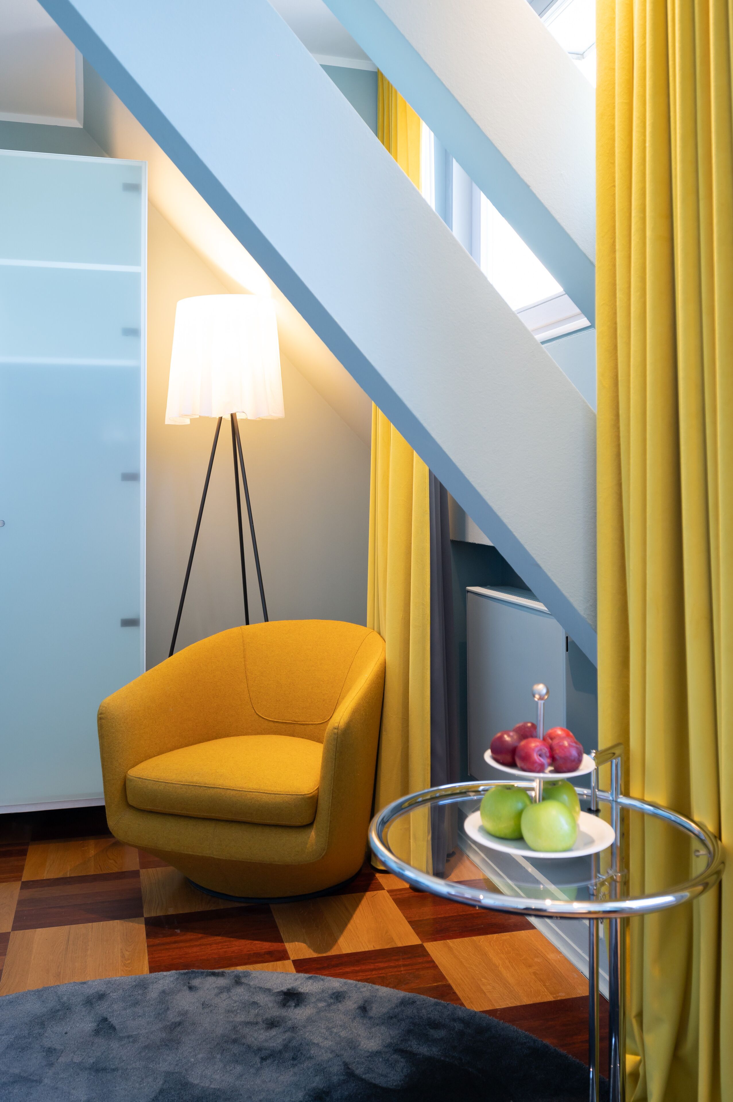 Uma sala de estar moderna com uma poltrona amarela, um abajur, uma mesa de vidro com frutas e uma escada com cortinas amarelas.