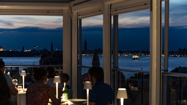 Um terraço com mesas e cadeiras, com vista para um corpo de água e luzes da cidade ao fundo.