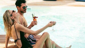 Um casal sentado à beira da piscina, com bebidas, deck de madeira e água cristalina.