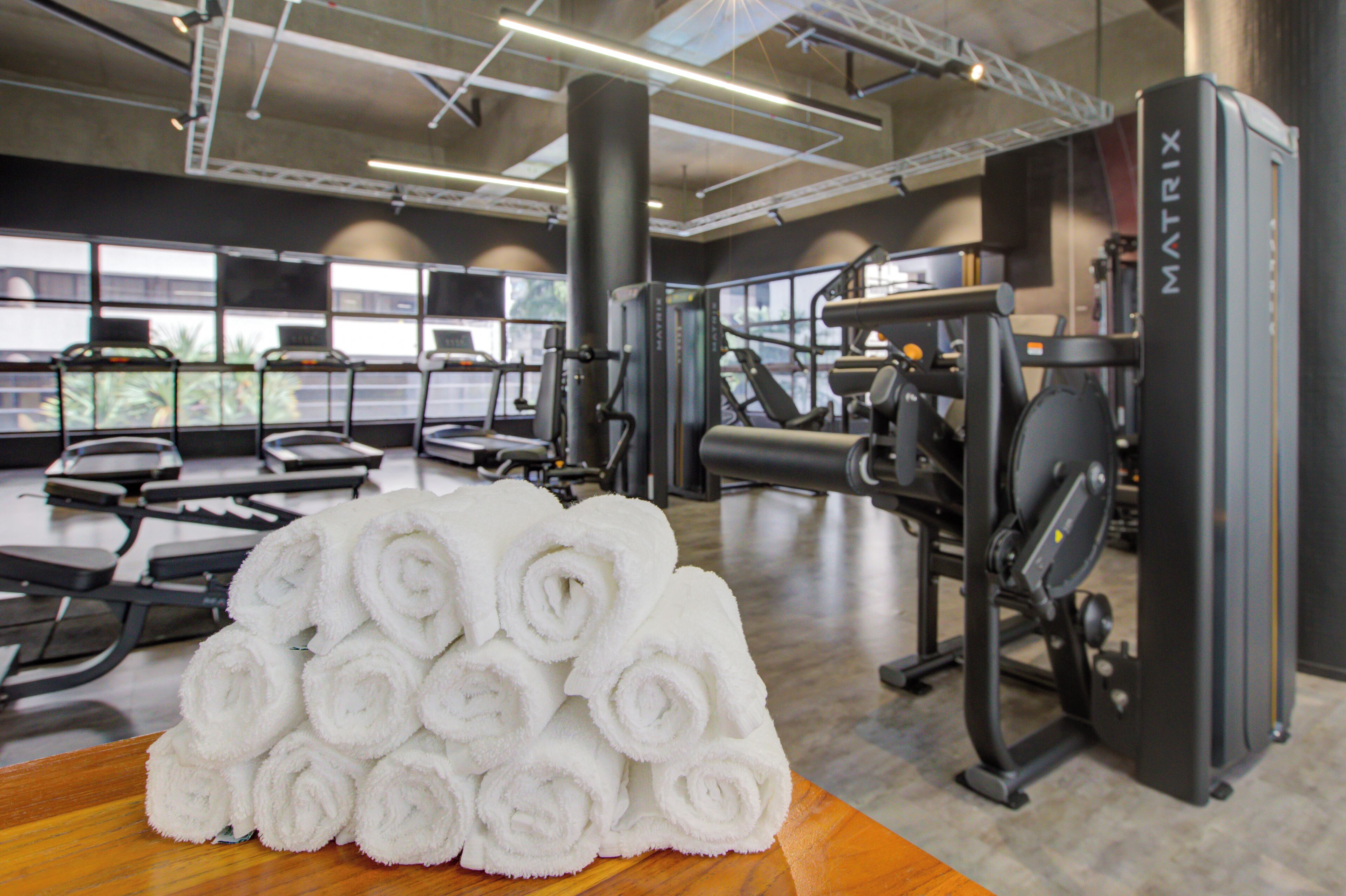 Uma academia com equipamentos de exercício, uma mesa de madeira com toalhas dobradas e amplas janelas.