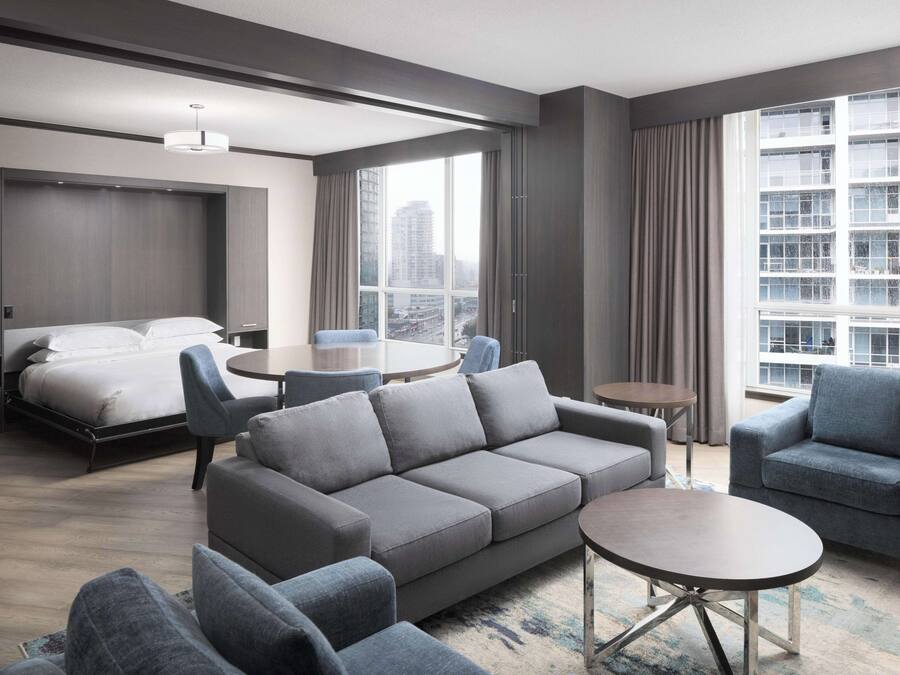 Quarto de hotel moderno com uma cama grande, sofá cinzento, poltronas, uma mesinha de centro e vista para a cidade através de grandes janelas.