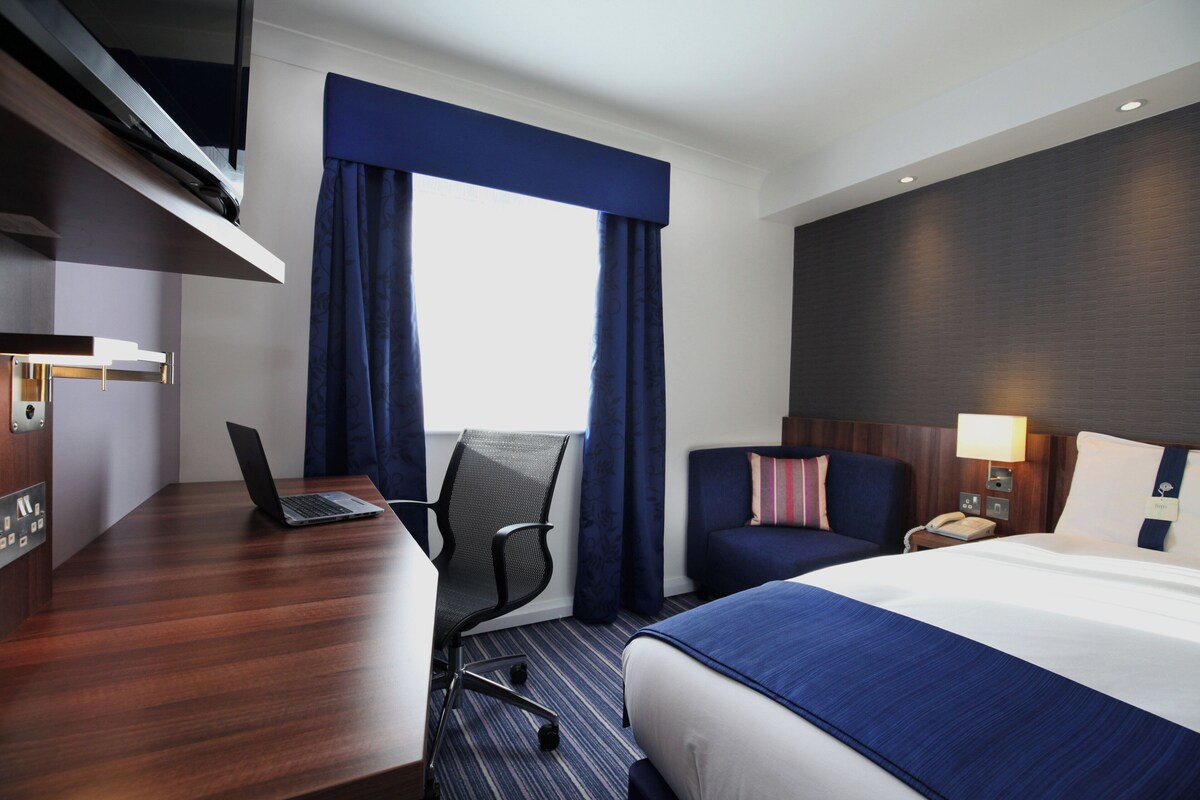Quarto de hotel com cama, escrivaninha com laptop, cadeira, mesa de cabeceira com luminária e janela com cortinas.