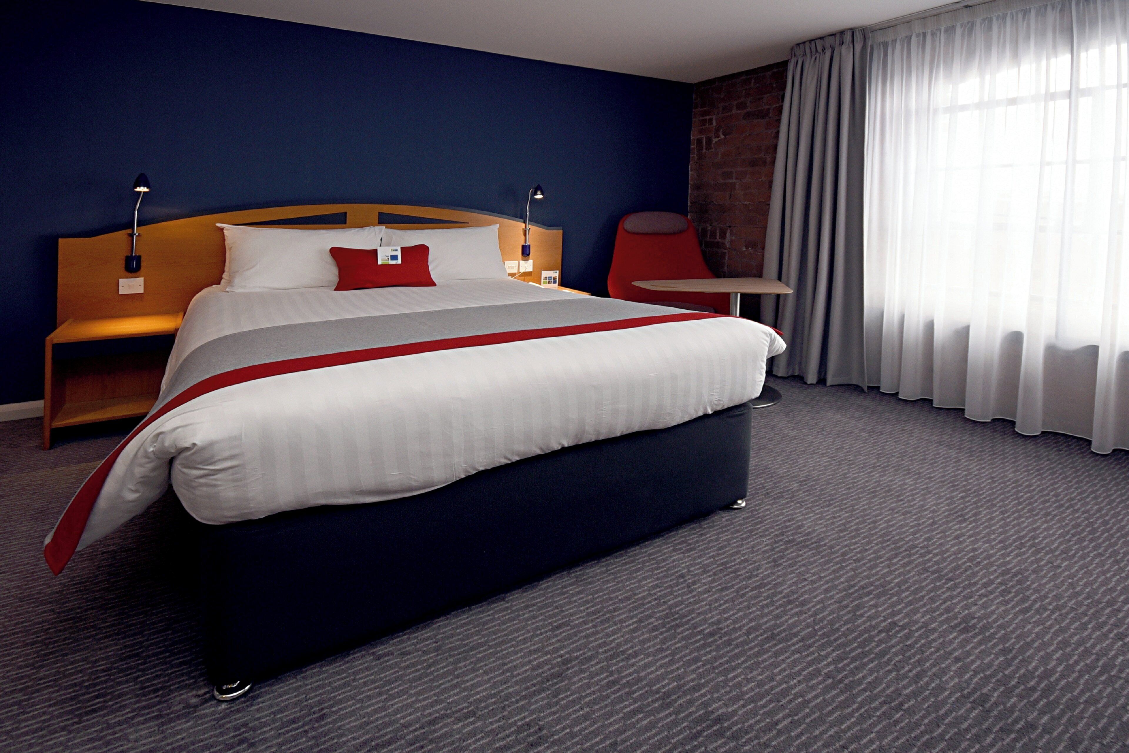 Quarto de hotel com uma cama grande, uma cadeira vermelha, cabeceira de madeira e parede de tijolos.