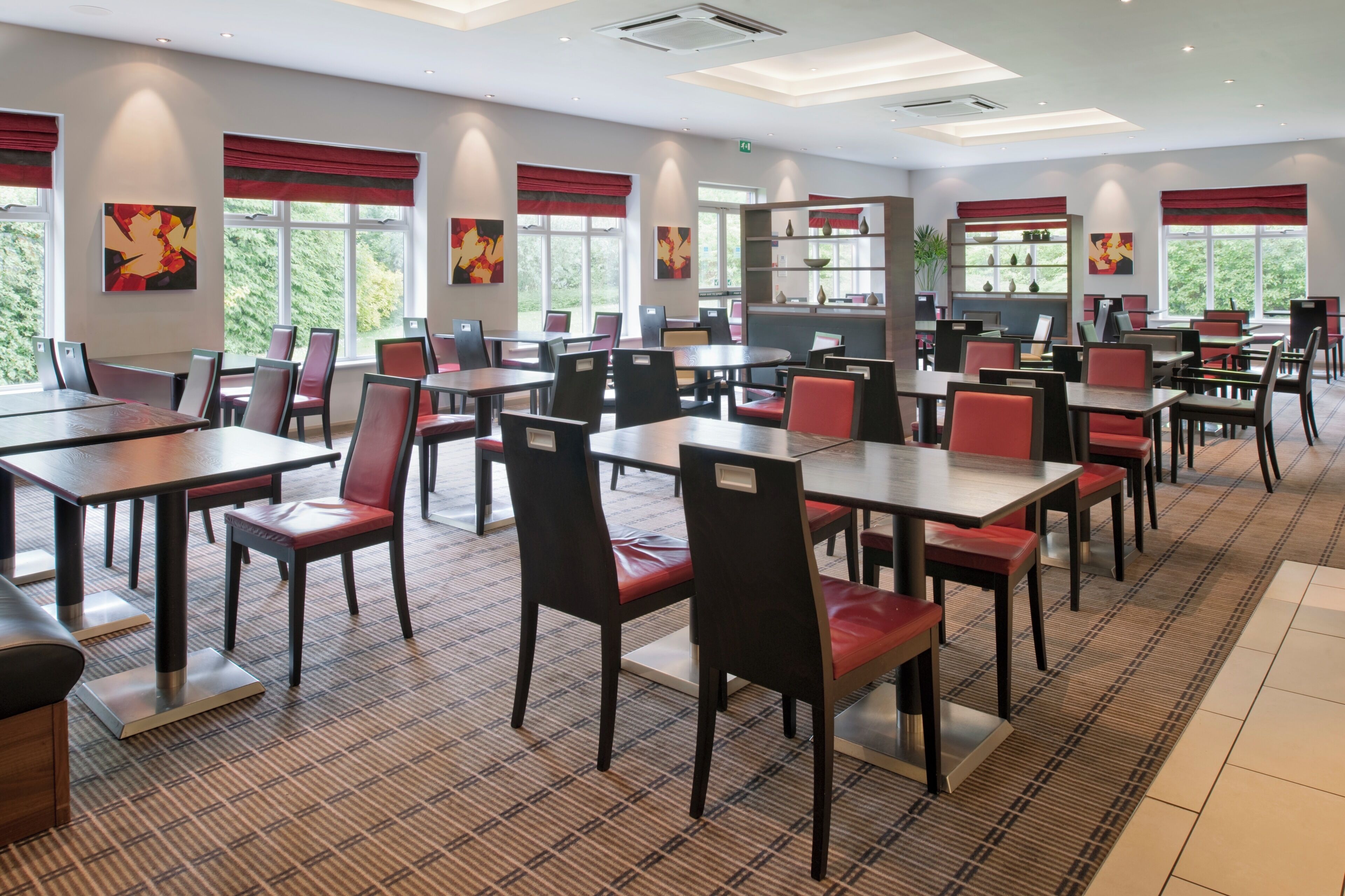 Um restaurante amplo com várias mesas e cadeiras, grandes janelas com cortinas vermelhas e obras de arte nas paredes.