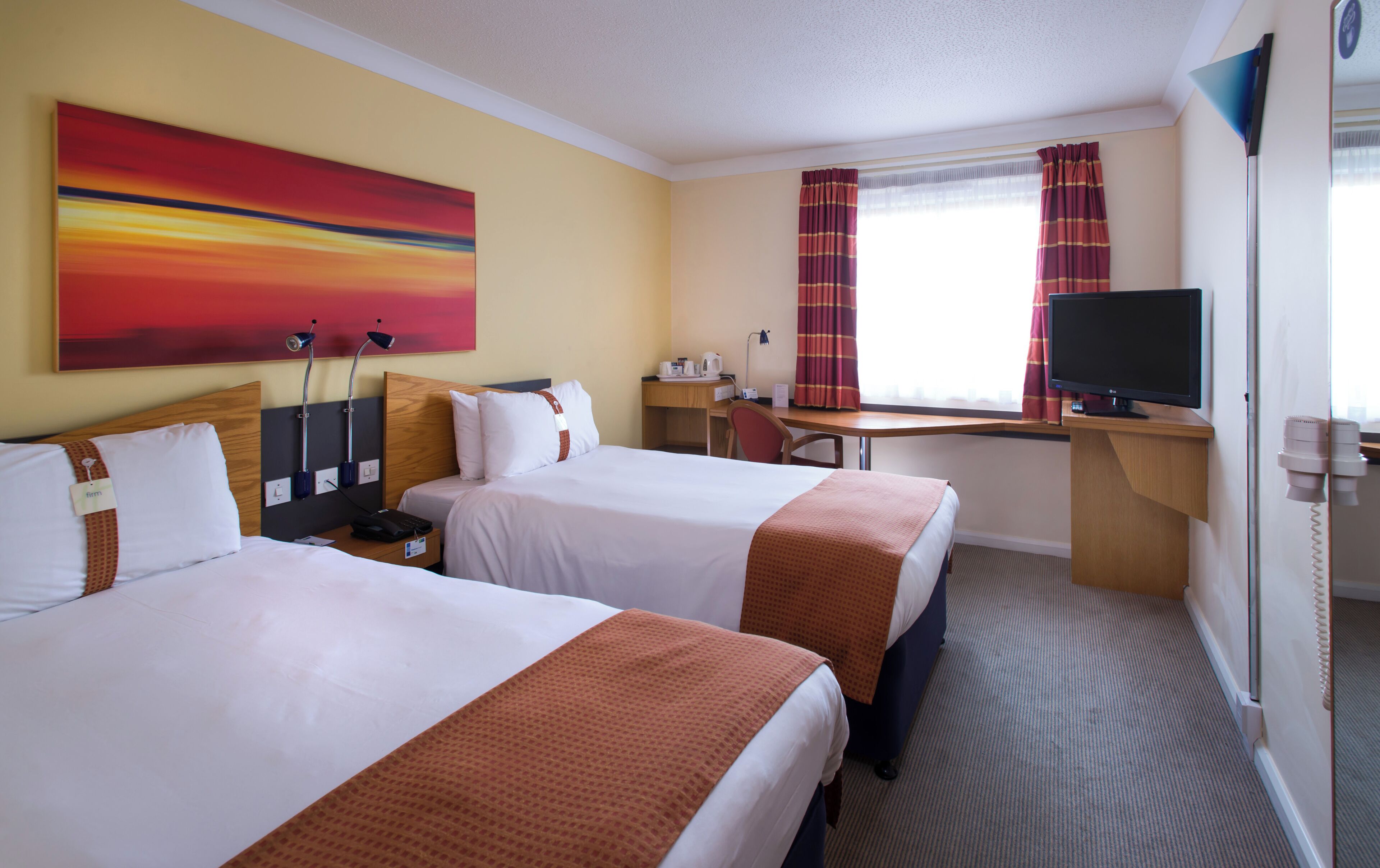 Quarto de hotel com duas camas, uma escrivaninha com computador, televisão e um quadro na parede.