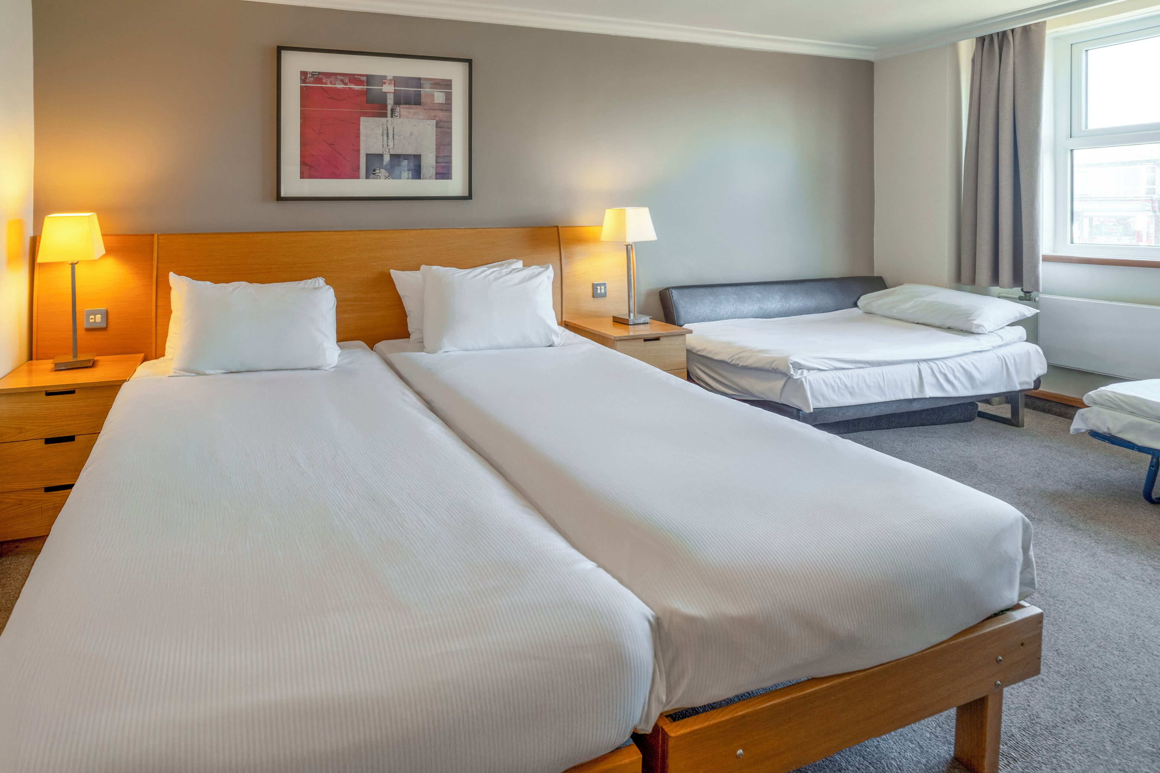 Quarto de hotel com duas camas, cabeceira de madeira, abajures, um quadro na parede e uma janela com cortinas.