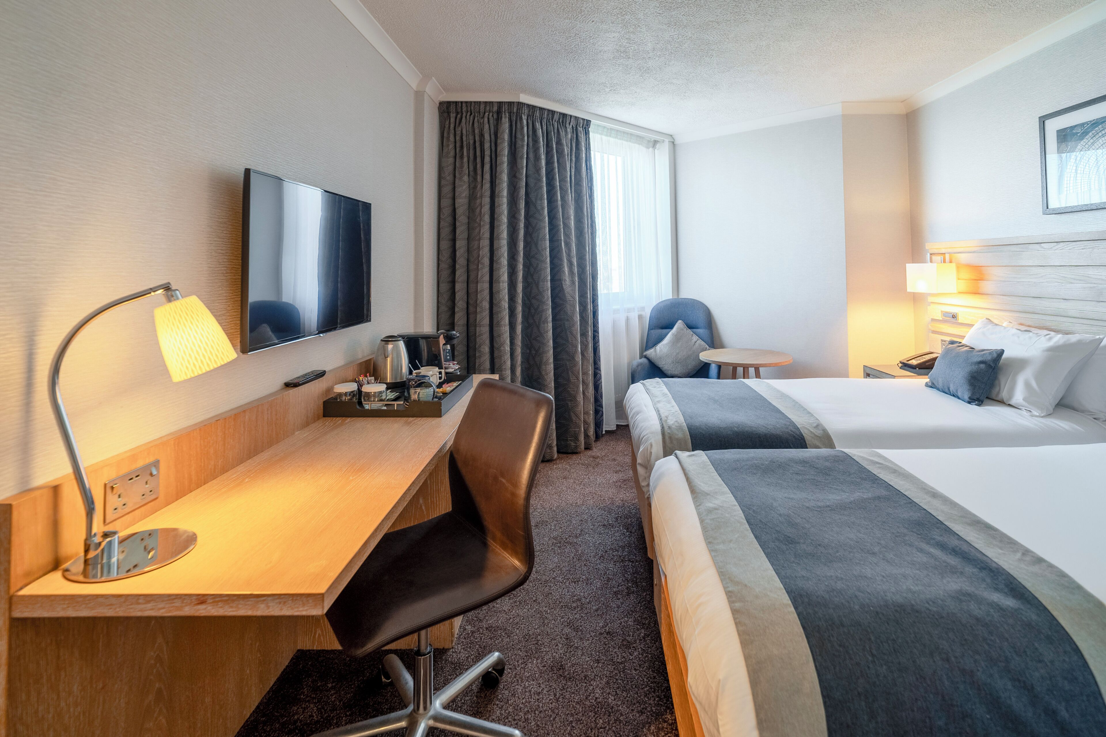 Quarto de hotel com escrivaninha, cadeira, duas camas, televisão, mesa pequena e janela com cortinas.