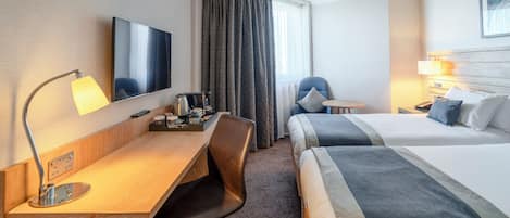 Quarto de hotel com escrivaninha, cadeira, duas camas, televisão, mesa pequena e janela com cortinas.