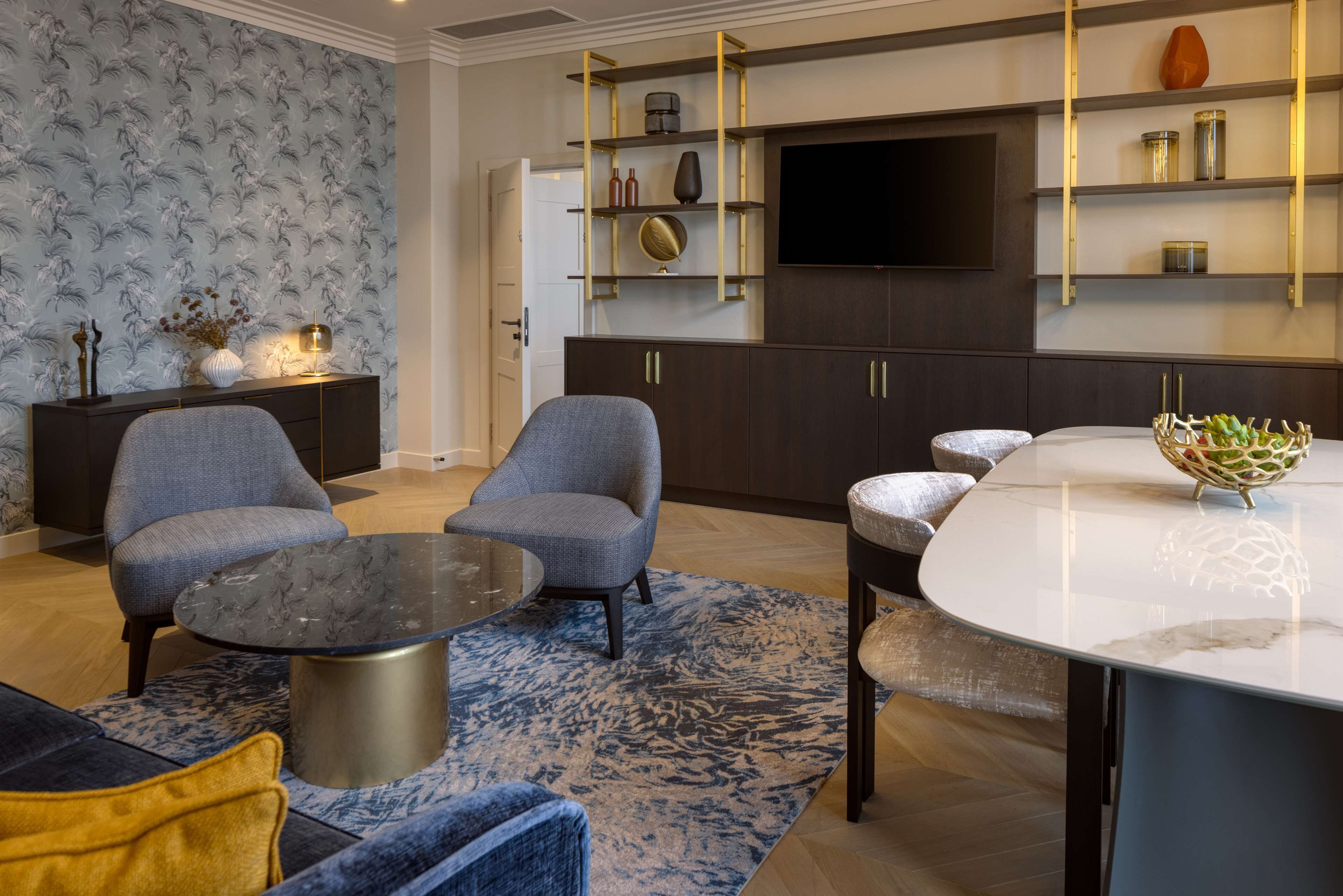 Uma sala de estar moderna com um sofá, poltronas, uma mesa de centro e uma mesa de jantar. Há uma TV fixada na parede, uma estante de livros e uma taça decorativa na mesa de jantar.