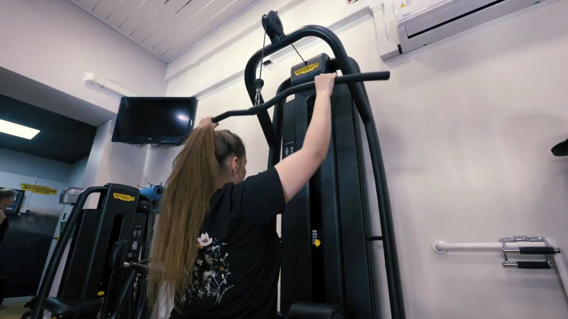 Uma academia com equipamentos de exercícios, uma televisão e um ar condicionado fixado na parede.