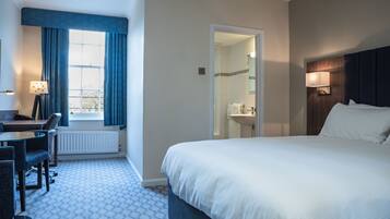 Quarto de hotel com uma cama grande, uma secretária com cadeira, uma janela com cortinas, um banheiro com espelho e pia, e uma luminária na parede.