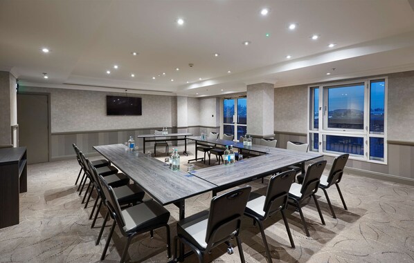Sala de conferências com uma mesa comprida, cadeiras, TV de tela plana e grandes janelas com vista para a cidade.