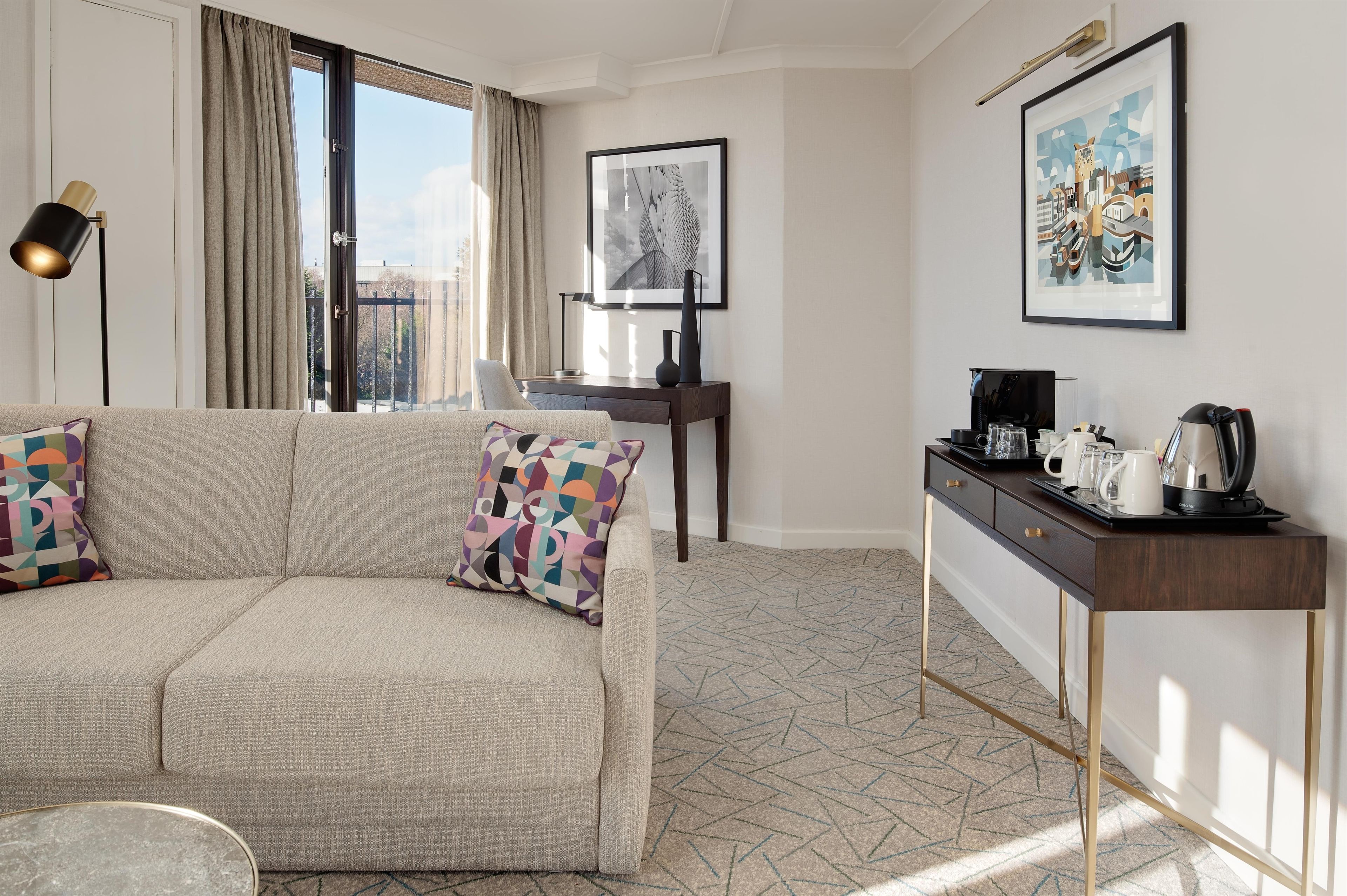 Quarto de hotel moderno com sofá, escrivaninha, cafeteira e obras de arte nas paredes.