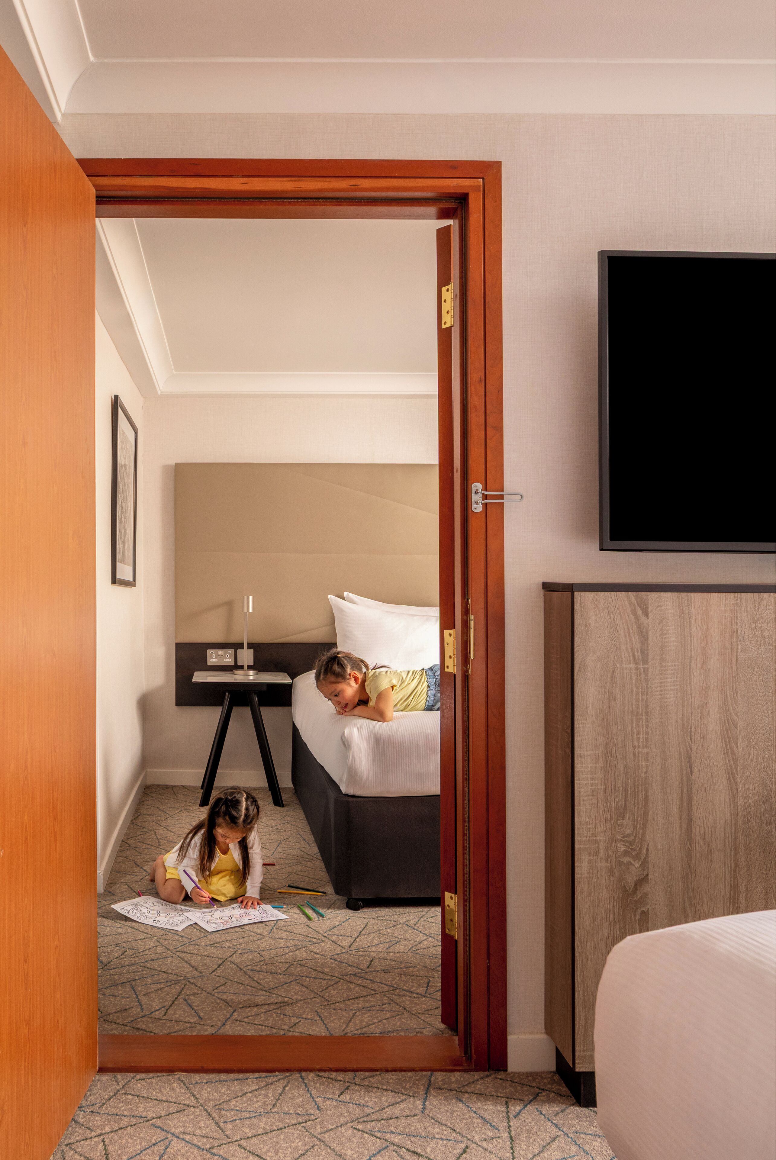 Quarto de hotel com cama, televisão, mesa de cabeceira e porta que dá para outro espaço.