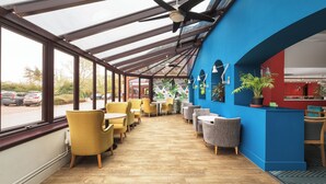 Um restaurante com uma longa mesa de madeira e cadeiras amarelas, parede azul com balcão vermelho e teto de vidro.