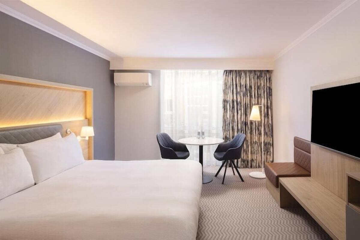 Quarto de hotel com cama, televisão, secretária com duas cadeiras e janela com cortinas.