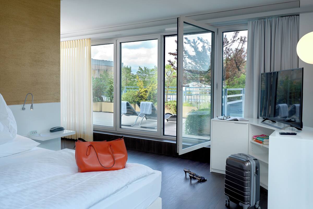 Quarto de hotel com cama, televisão, escrivaninha com controle remoto, mala e janela com vista para o exterior.