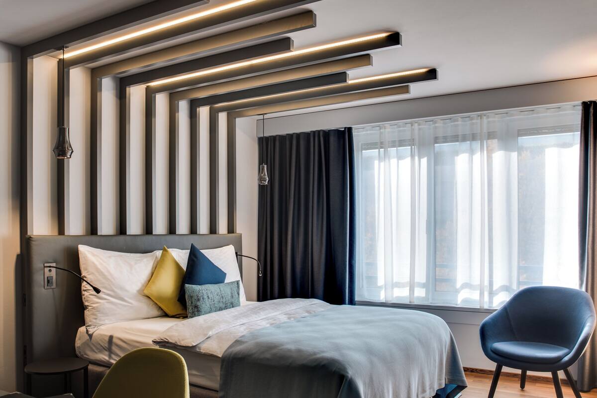 Quarto de hotel moderno com cama, mesa de cabeceira, cadeira e uma janela grande com cortinas.