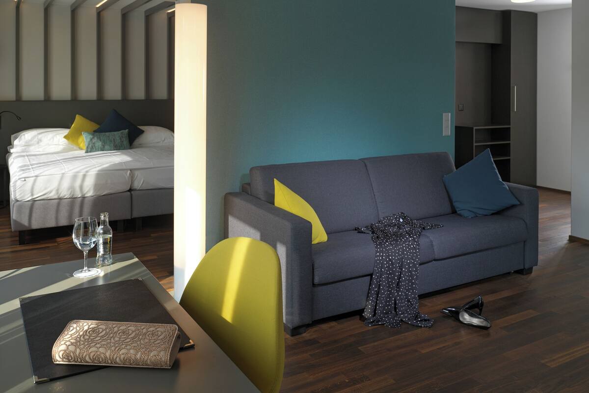 Quarto de hotel moderno com cama, sofá cinza e área de refeições com mesa e cadeiras.