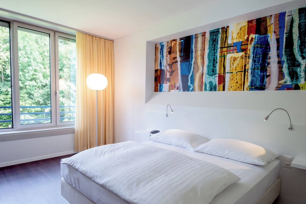 Quarto de hotel moderno com uma cama grande, um quadro na parede, uma janela com cortinas e um abajur.