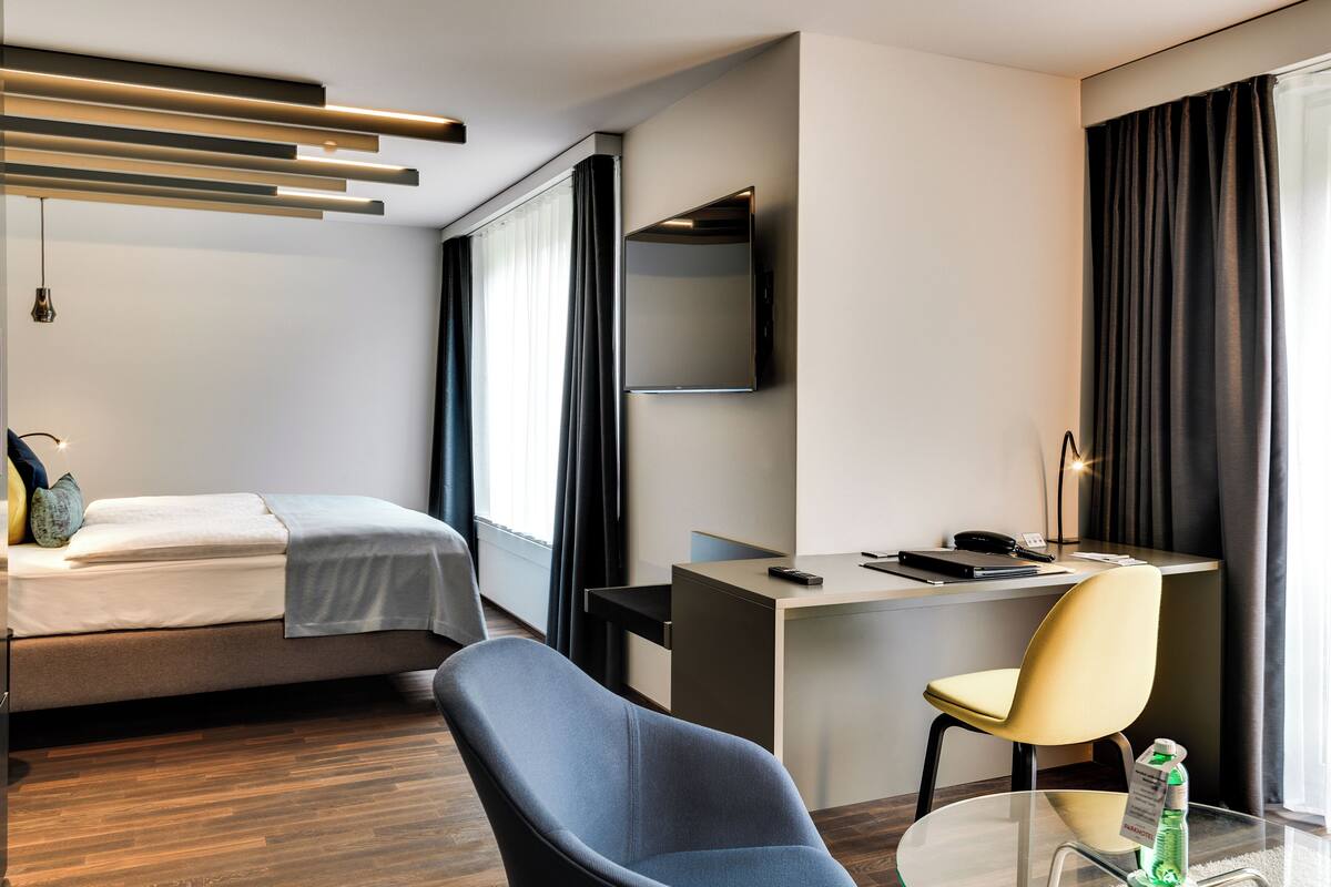Quarto de hotel moderno com cama, escrivaninha, cadeira e mesa de vidro.
