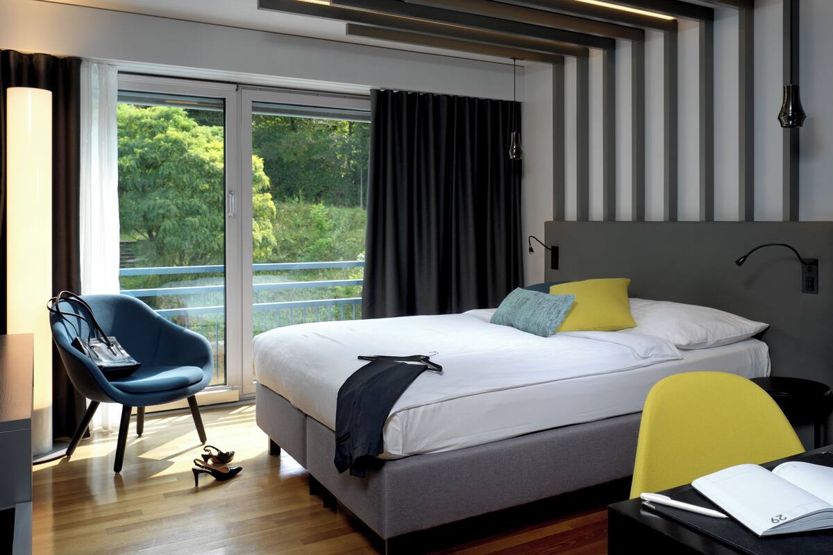 Quarto de hotel moderno com uma cama grande, uma escrivaninha, uma cadeira e vista para o verde lá fora.