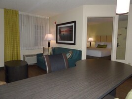 Quarto de hotel com mesa de jantar de madeira, sofá azul, cama com roupa de cama branca e um quadro na parede.