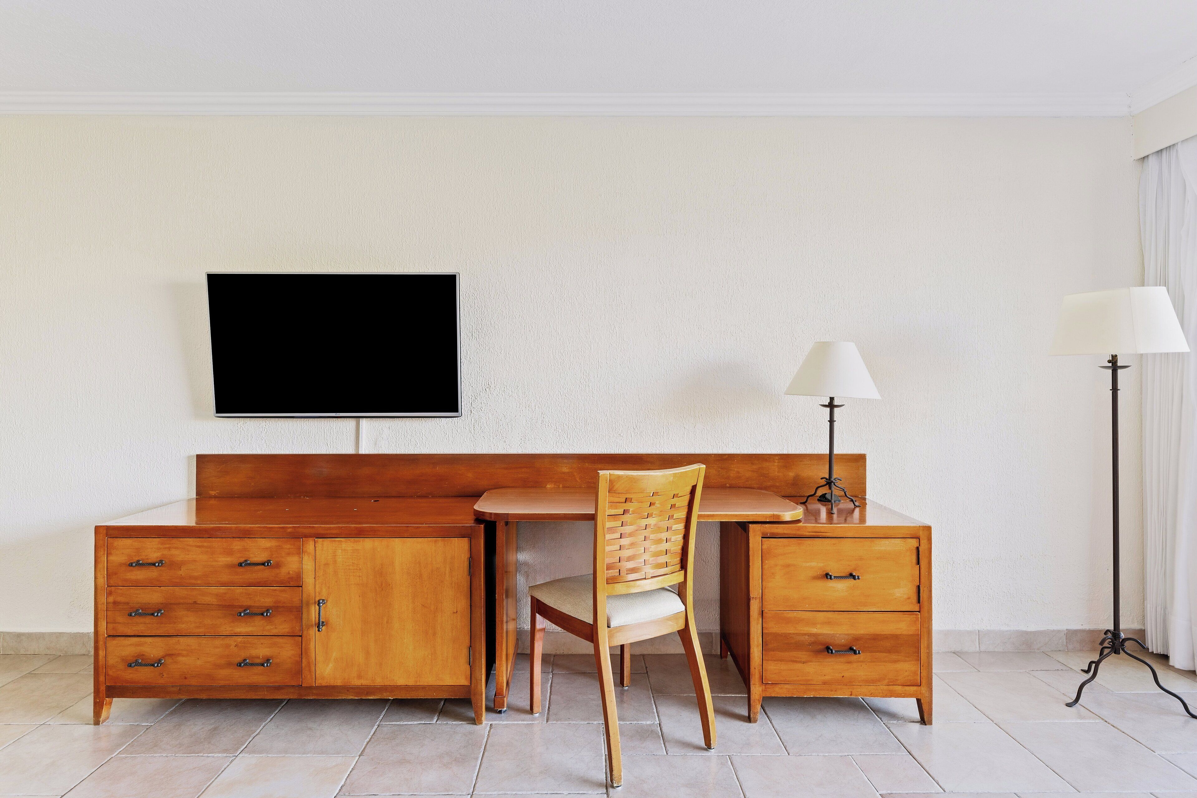 Uma mesa de madeira com gavetas, uma cadeira, uma televisão fixada na parede e um abajur.