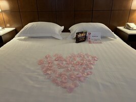 Quarto de hotel com uma cama bem arrumada, um arranjo de pétalas de rosa em formato de coração, um minibar e um sinal oferecendo chocolate grátis.