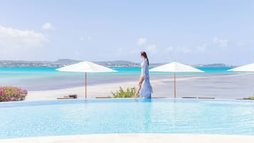 Uma mulher de vestido listrado caminhando à beira da piscina, com guarda-sóis e vista para a praia ao fundo.