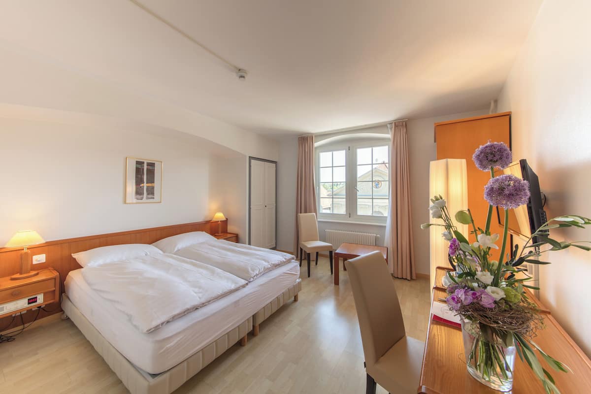 Quarto de hotel com cama de casal, abajures, escrivaninha com cadeira, vaso com flores e janela com cortinas.