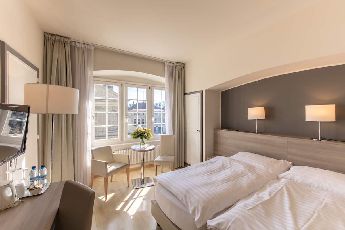 Quarto de hotel com cama, abajures, escrivaninha com luminária, cadeira, mesinha com um vaso e janela com cortinas.