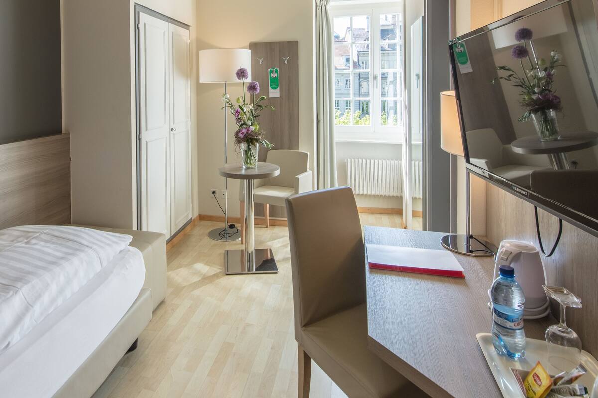 Quarto de hotel com cama, escrivaninha, cadeira, luminária, televisão e janela com vista para edifícios.