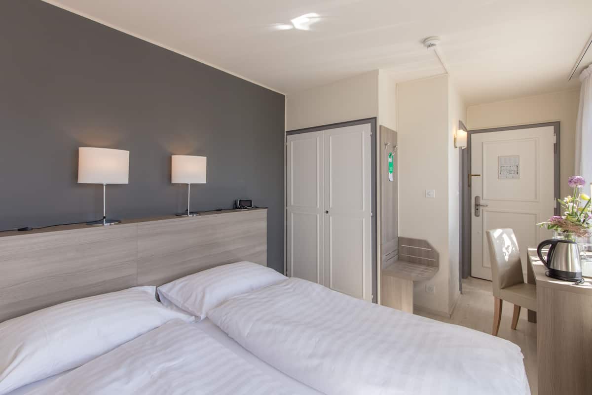 Quarto de hotel com cama de casal, duas luminárias de cabeceira, painel de cabeceira de madeira, guarda-roupa, escrivaninha com chaleira e uma cadeira.