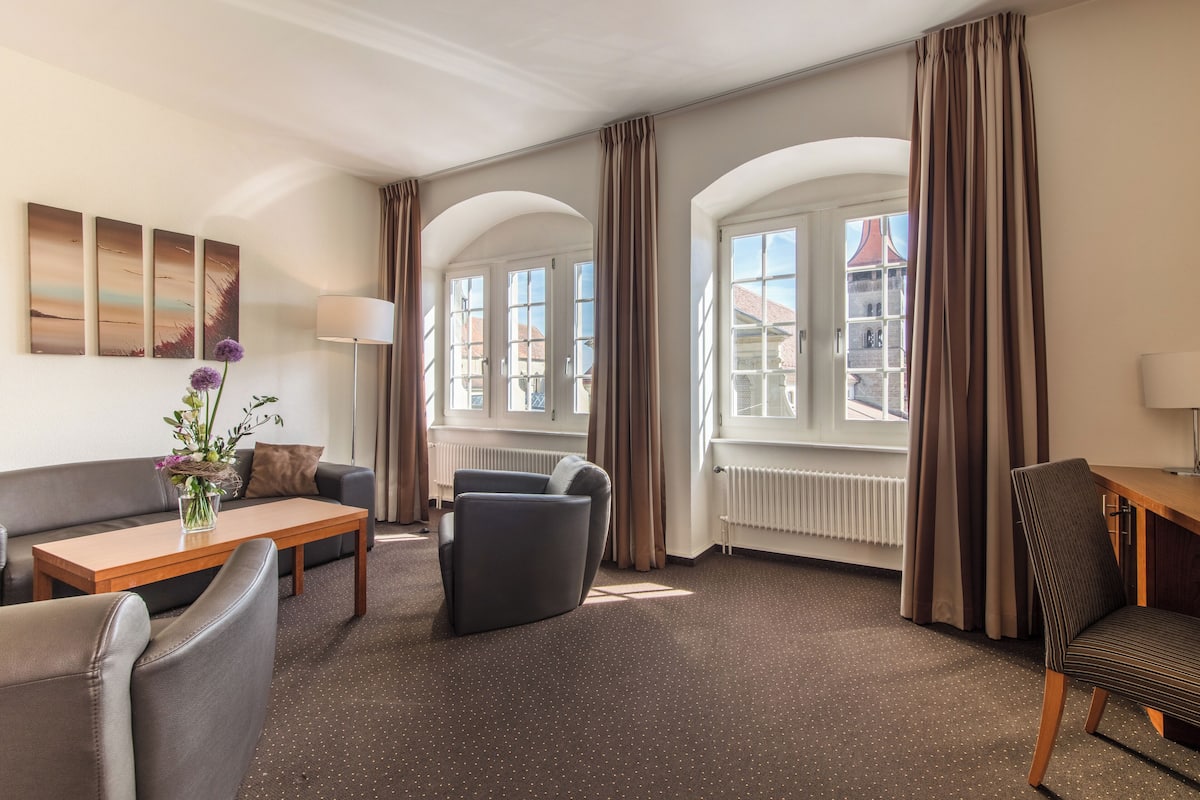 Quarto de hotel moderno com sofá, poltronas, uma mesa de madeira e vista para a cidade através de grandes janelas.