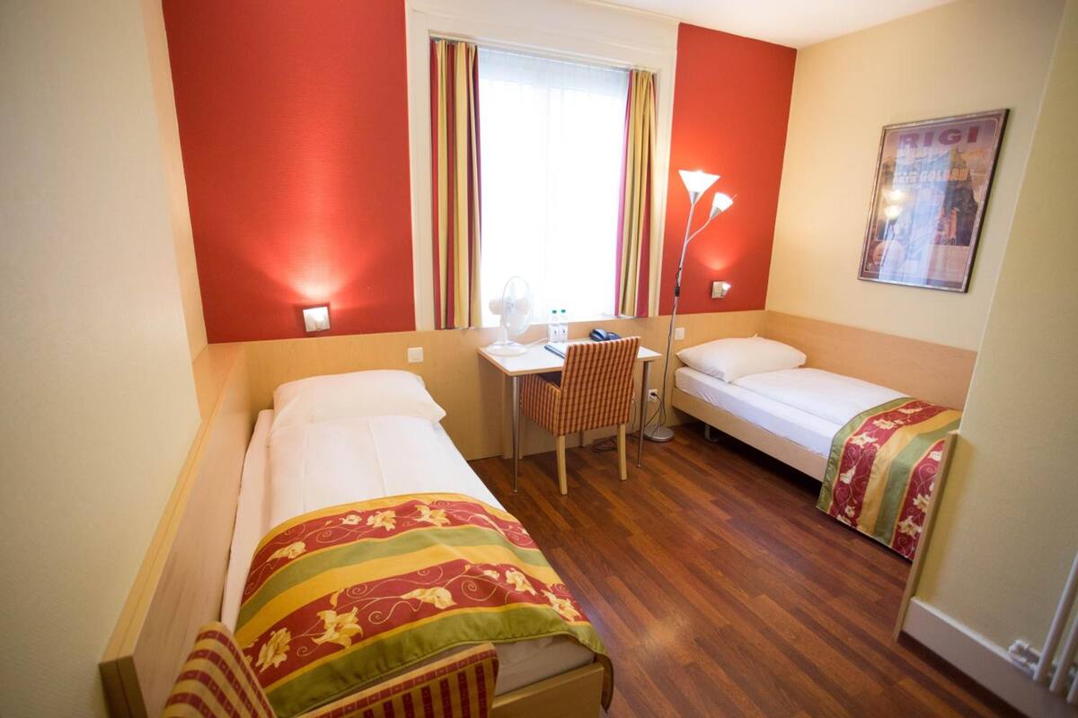Quarto de hotel com duas camas de solteiro, uma escrivaninha, uma cadeira, um abajur e um pôster na parede.