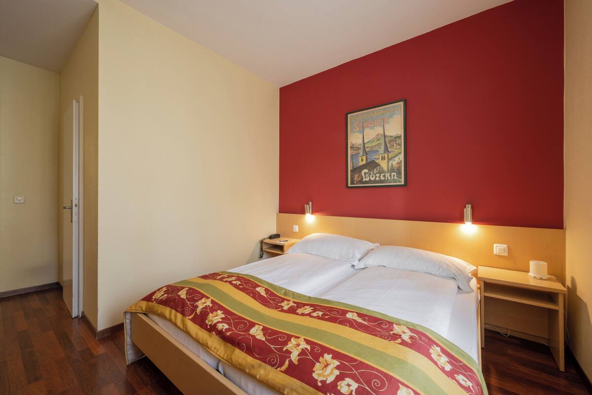 Quarto de hotel com uma cama, mesa de cabeceira, luminária e um quadro na parede retratando uma cidade.