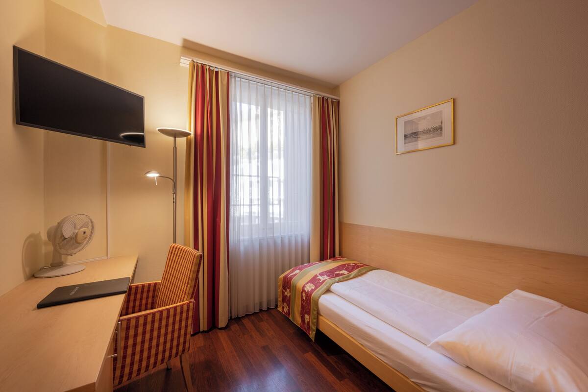 Quarto de hotel com uma cama, uma escrivaninha com um laptop, uma cadeira, um ventilador e uma janela com cortinas.