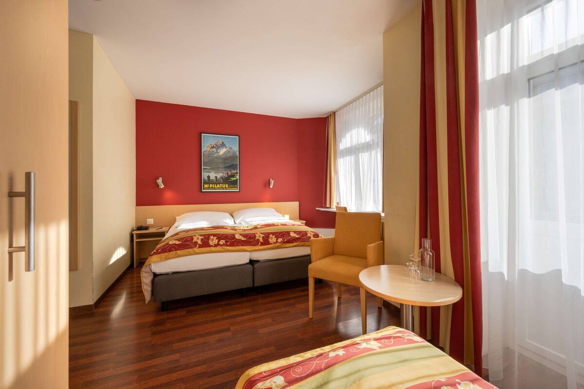 Quarto de hotel com uma cama, uma cadeira, uma mesa pequena e um cartaz na parede.