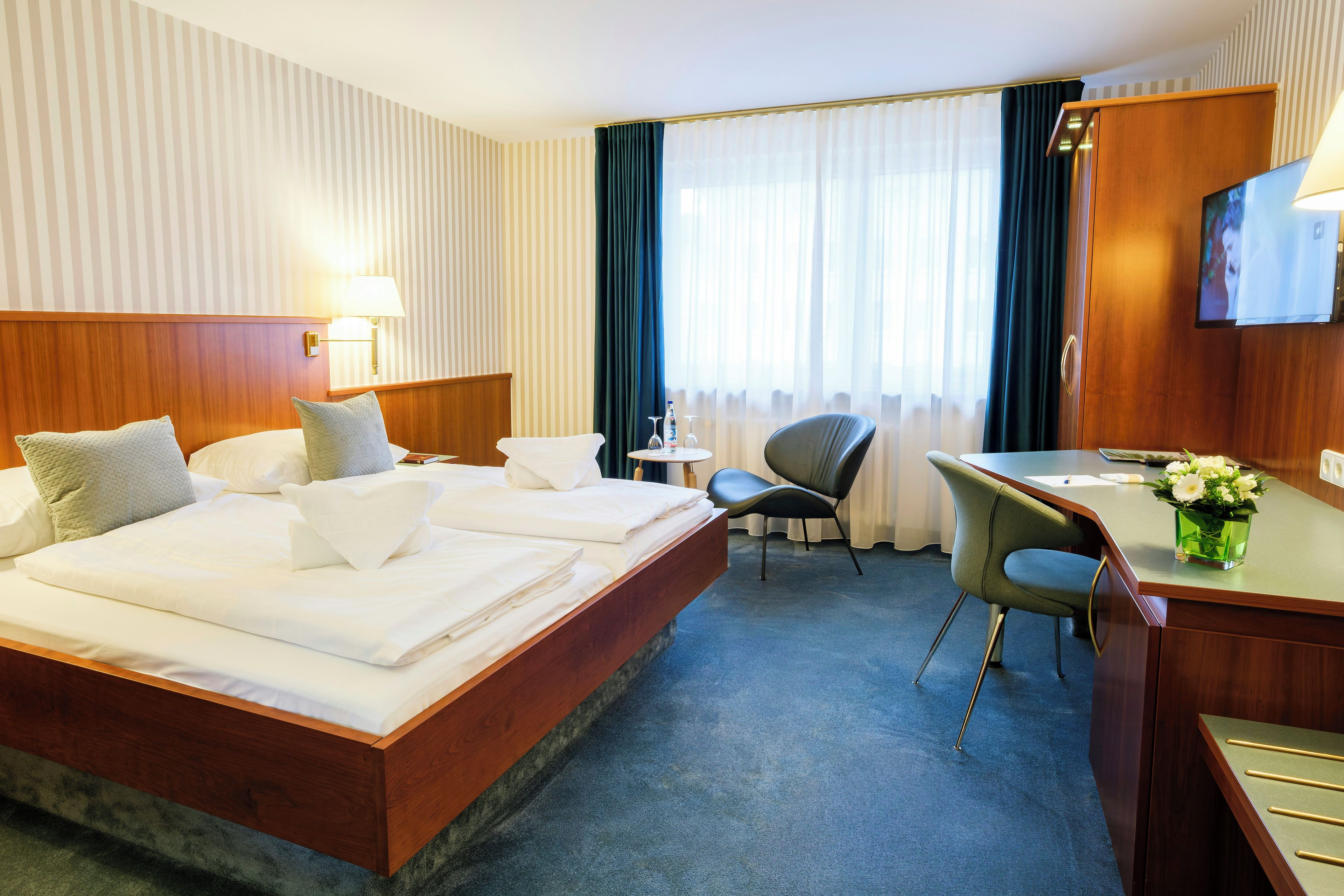 Quarto de hotel com cama grande, escrivaninha, cadeira, televisão e janela com cortinas.