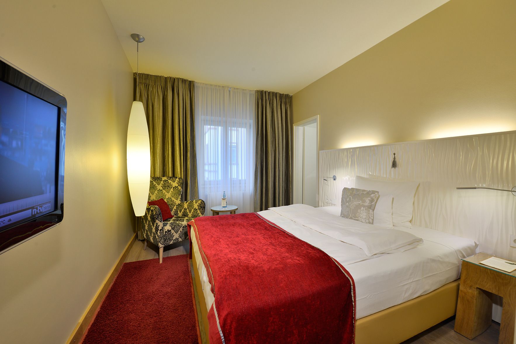 Quarto de hotel com cama, televisão, cadeira, mesa pequena e mesa de cabeceira.